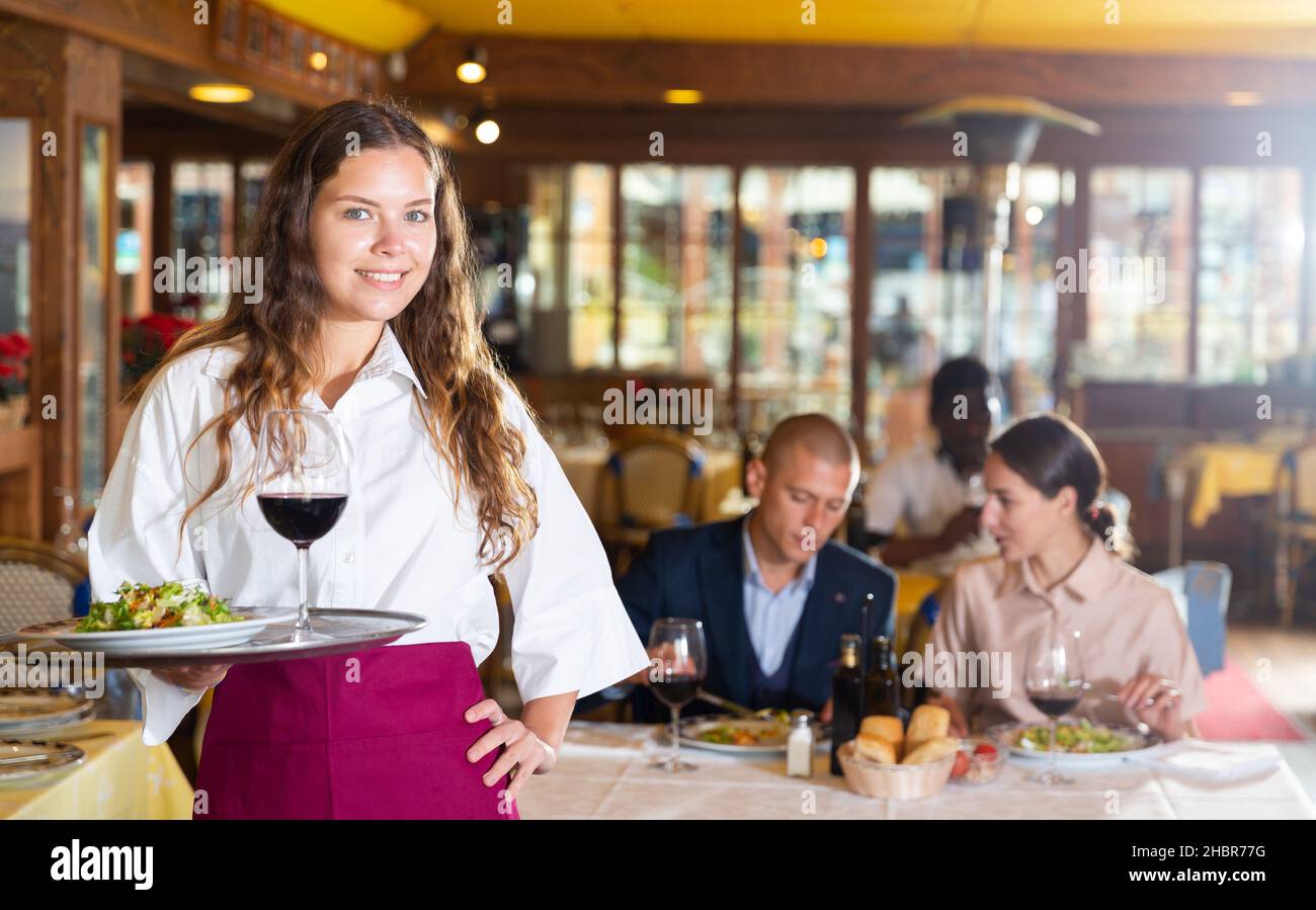 Successful young waitress standing in restaurant with ordered meals ...
