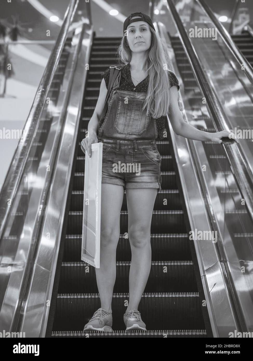 A girl in short denim overalls rides down the escalator holding a ...