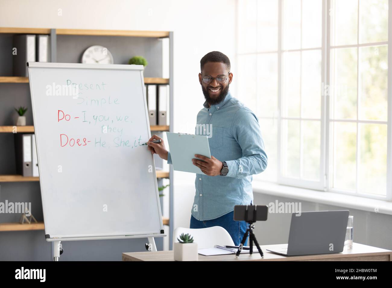 Happy young black man teacher in glasses looks at phone webcam shows to ...