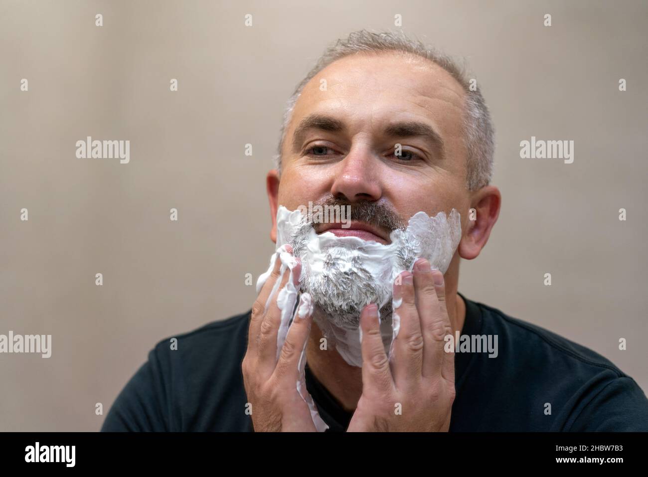 Portrait of handsome white-haired beared middle-aged man applying shaving foam to trim his beard ...
