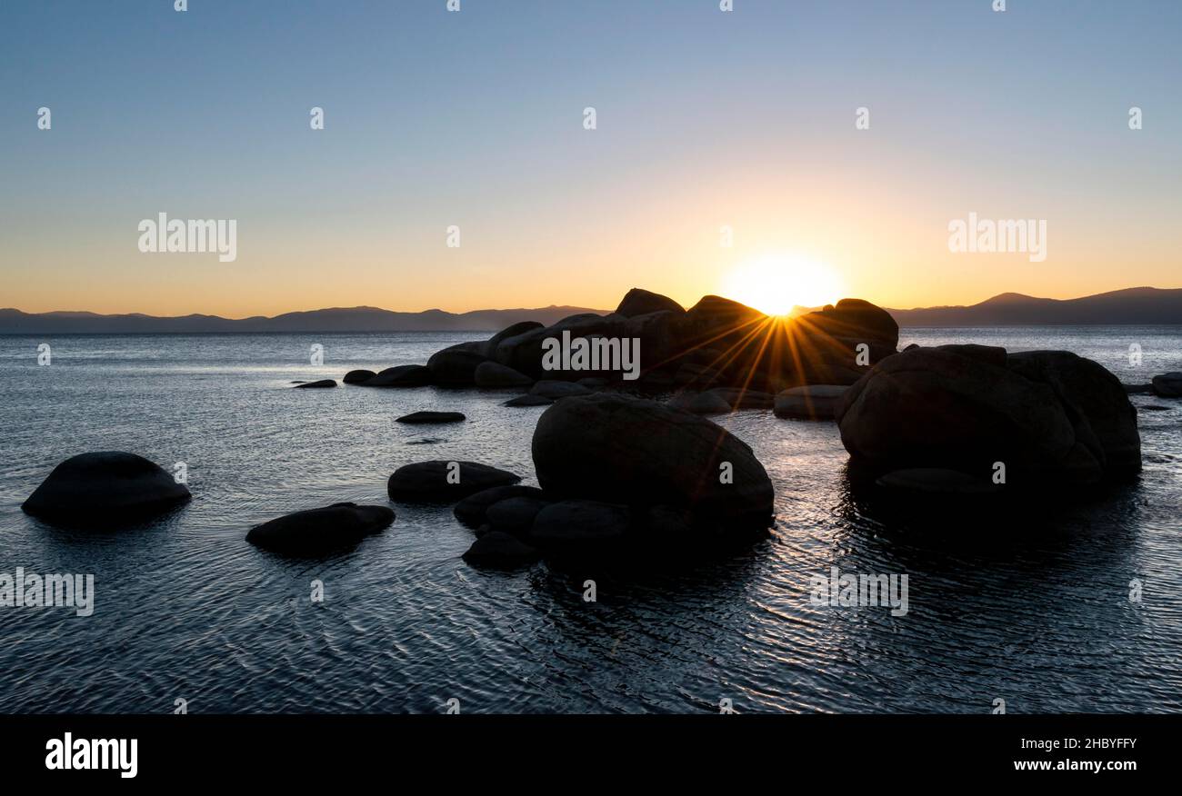 Sunset with sun star, round stones in the water, shore at Lake Tahoe ...