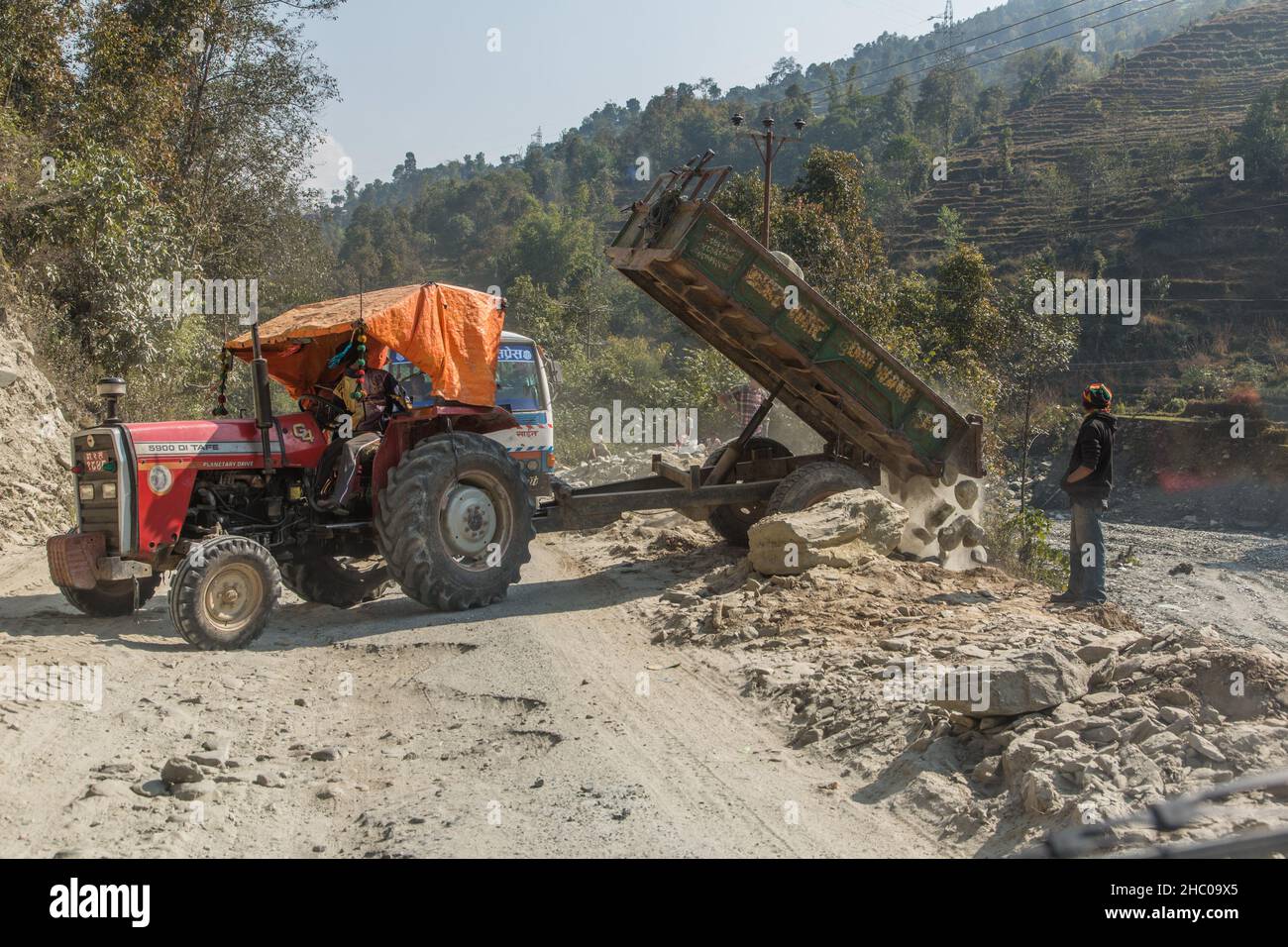 A tractor with a dump-trailer dumping a load of rocks to widen a road ...
