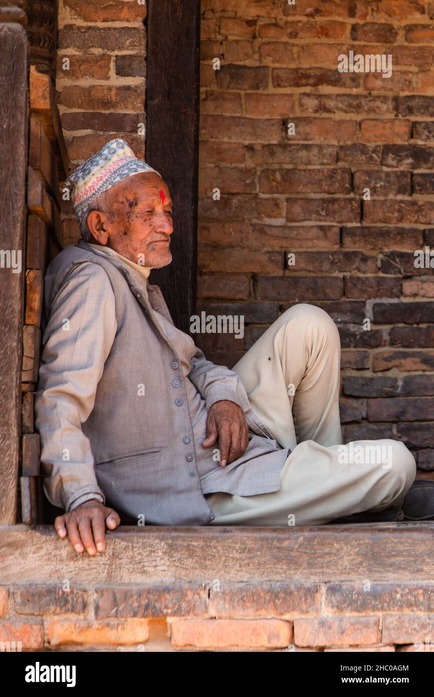A Nepali man in traditional attire and Dhaka topi cap sits in the shade ...