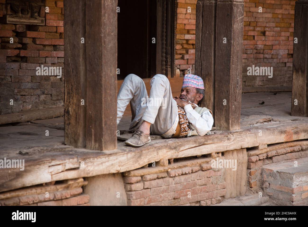 A Nepali man in traditional attire and Dhaka topi cap naps in the shade ...