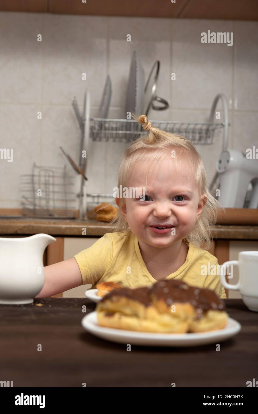 Cheerful blonde child in kitchen is having breakfast and looking at the ...