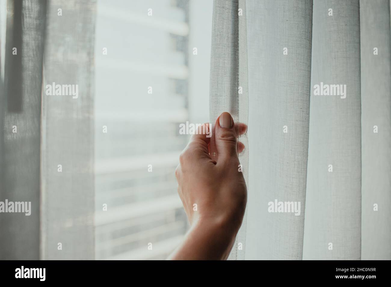 Person touching and holding a white curtain in room next to the window ...