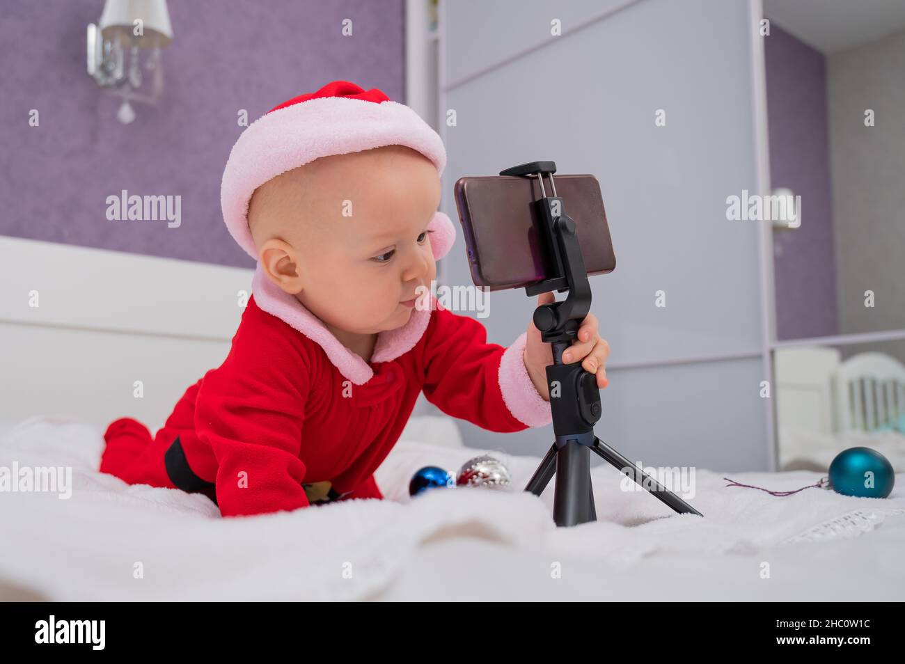A baby boy dressed in a Santa suit sits on the bed and looks at the ...