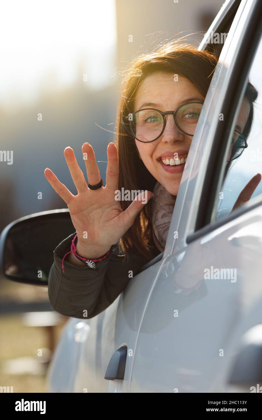 Young woman sat in car waving out out of the window with sunlight and ...
