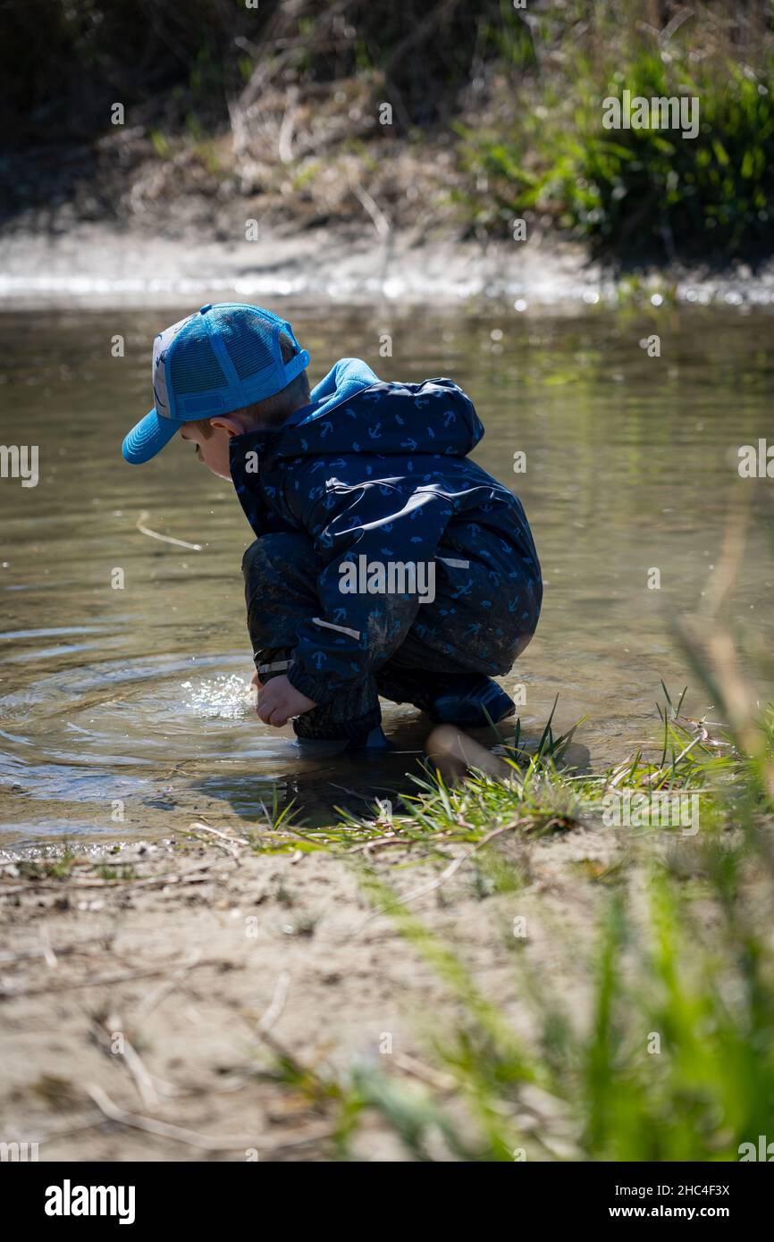 A young boy in a blue raincoat and rain boots happily splashes in a ...