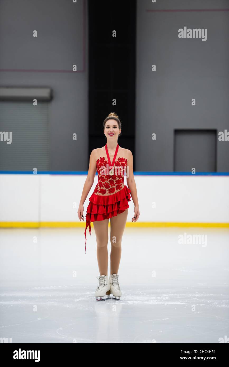 full length of cheerful figure skater in red dress with golden medal ...