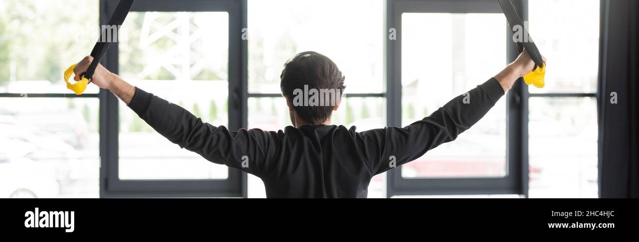Back view of sportsman working out with suspension straps in gym ...