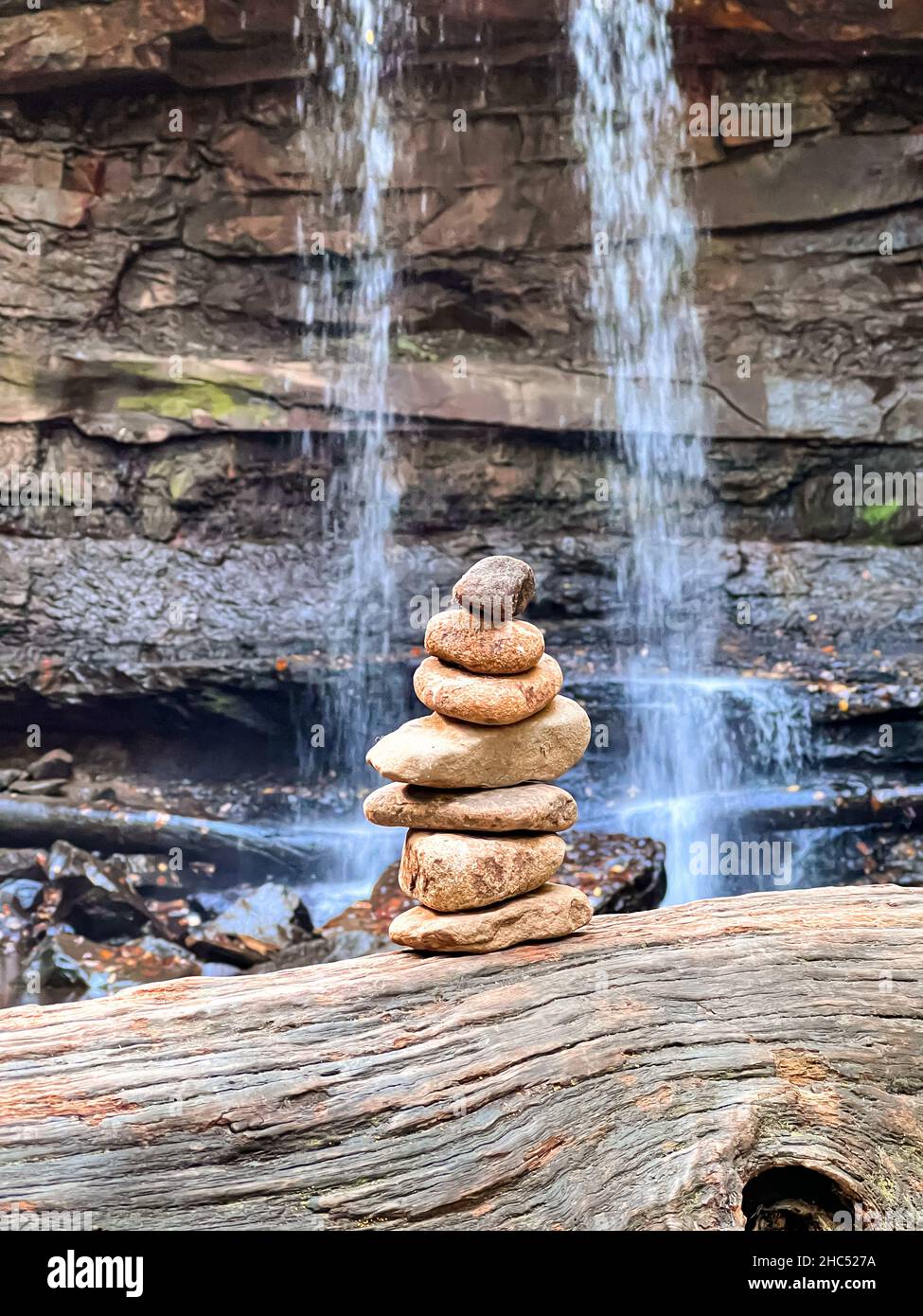 A vertical shot of a stack of stones on a log by the waterfall in a ...