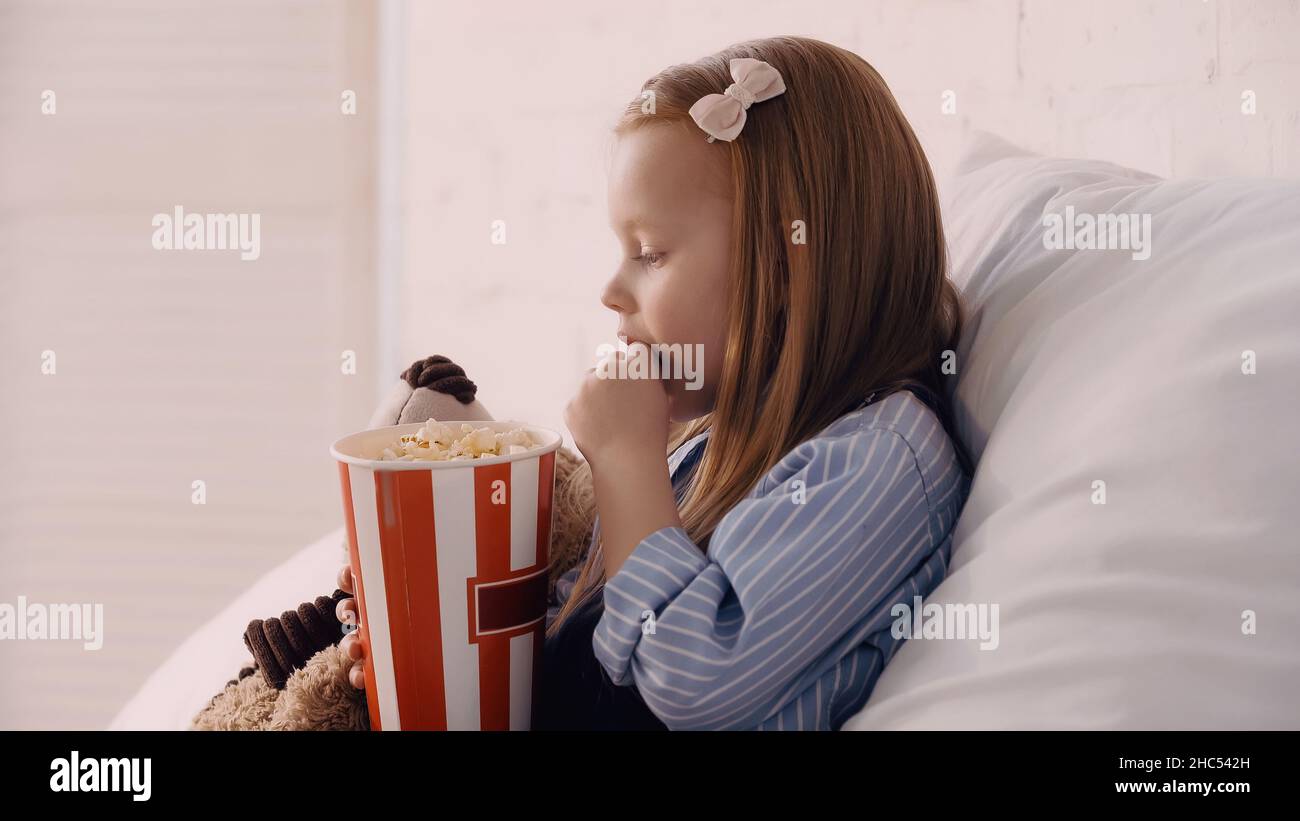 Side view of kid eating popcorn near toy on bed Stock Photo - Alamy