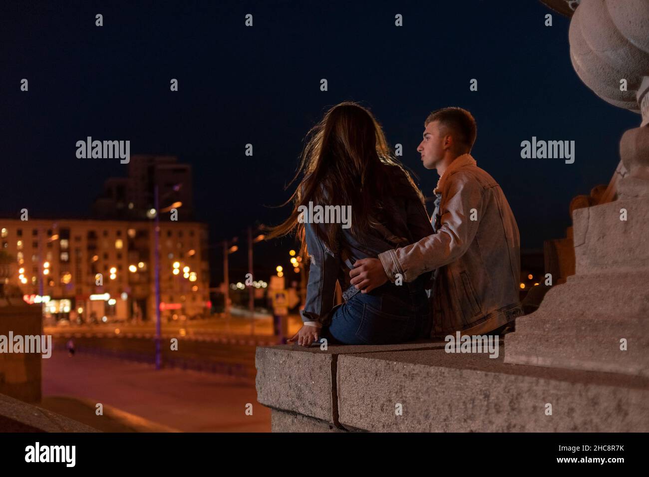 Young couple in love hugs sitting on a tall parapet over the evening ...