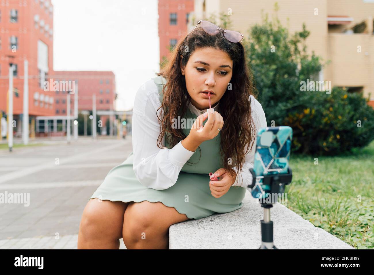 Young curvy influencer recording on smart phone while applying lipstick ...