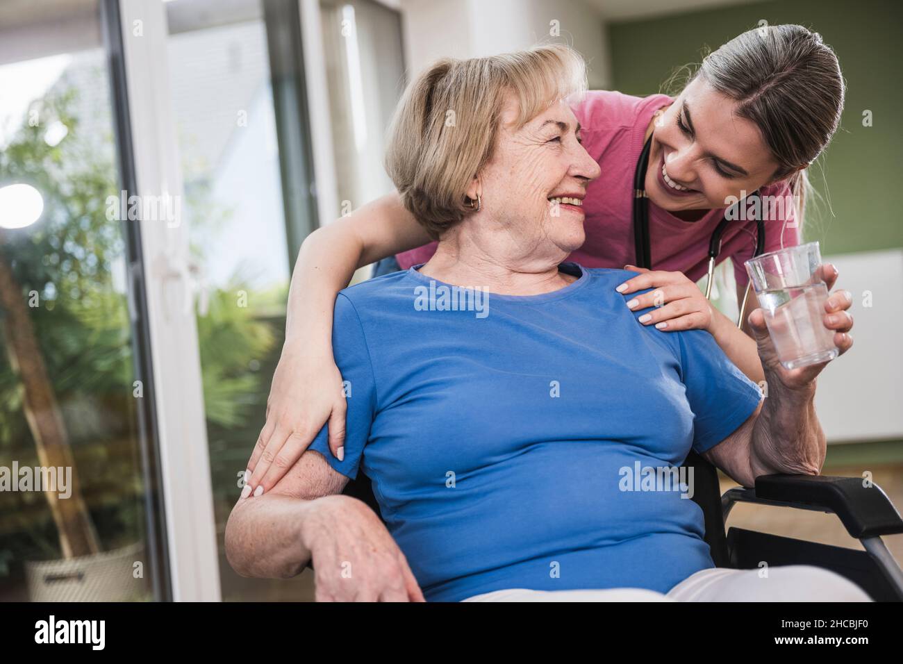 Smiling caregiver standing behind disabled woman in wheelchair at home ...