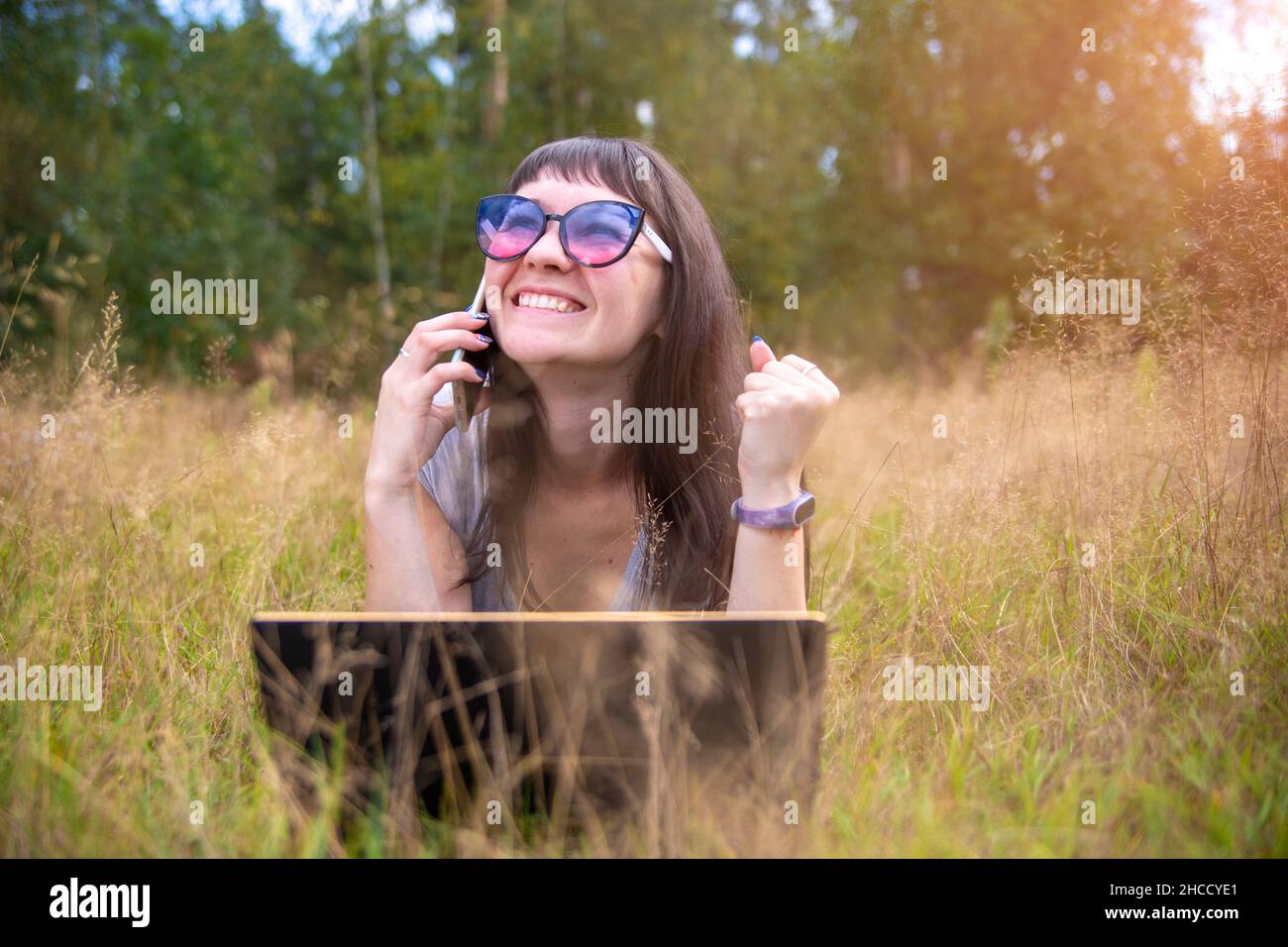 young woman smiling happily calling by phone with laptop on summer lawn ...
