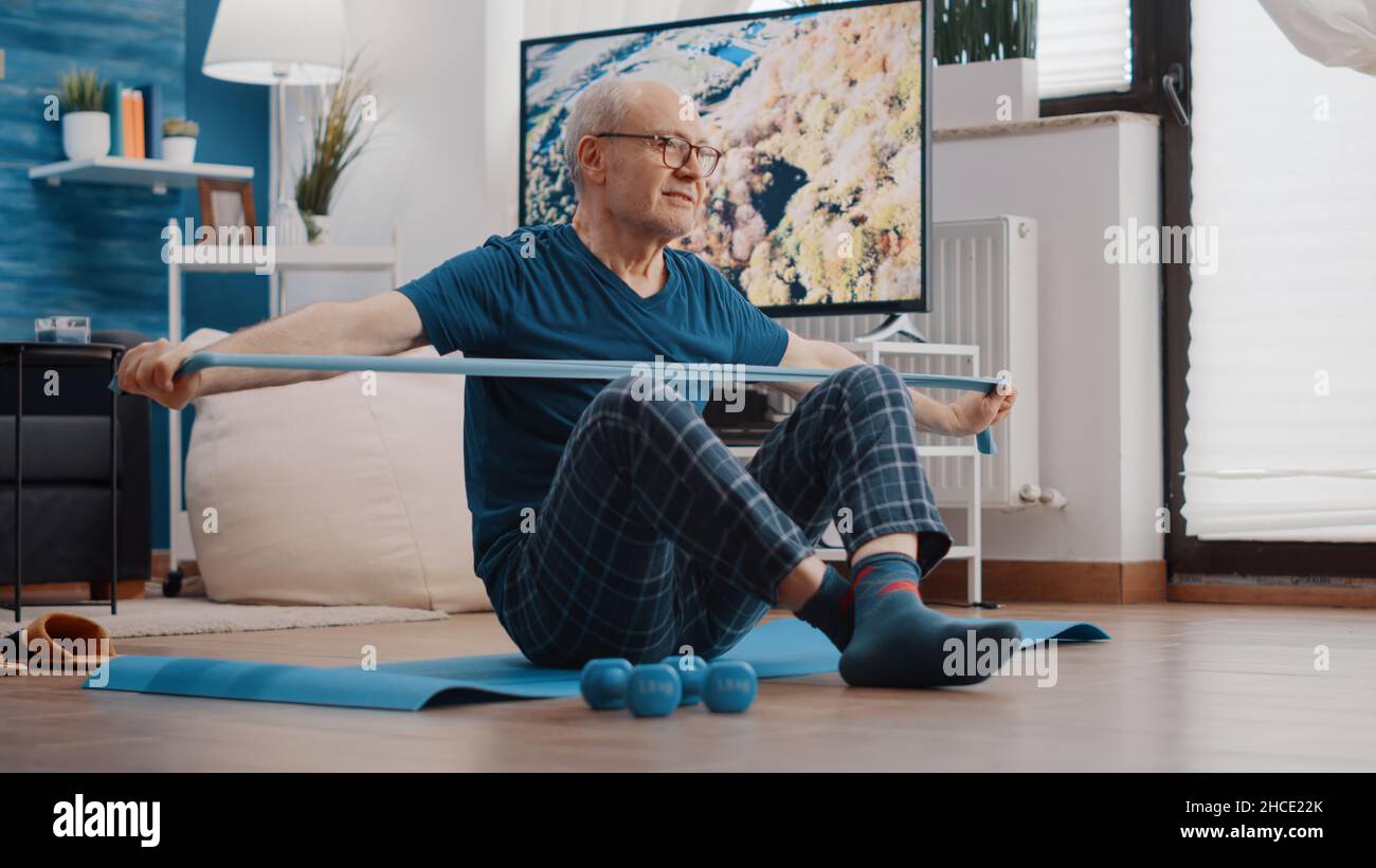 Old man using resistance band to stretch and pull, sitting on yoga mat ...