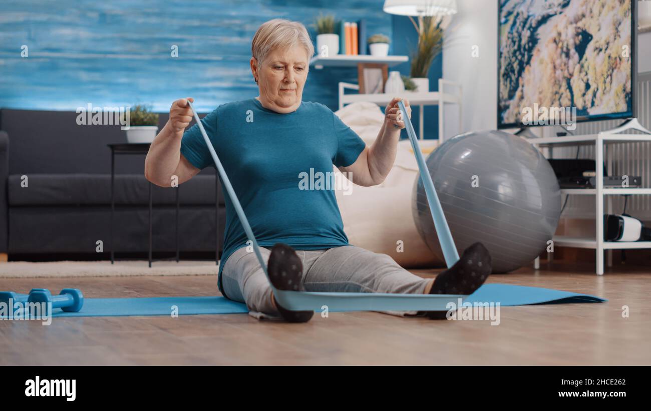 Aged woman using resistance band to train muscles on yoga mat. Retired ...