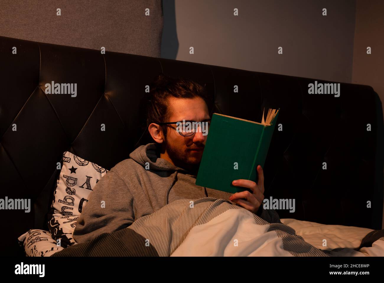 A young man laying in bed reading a book before going to sleep, after ...