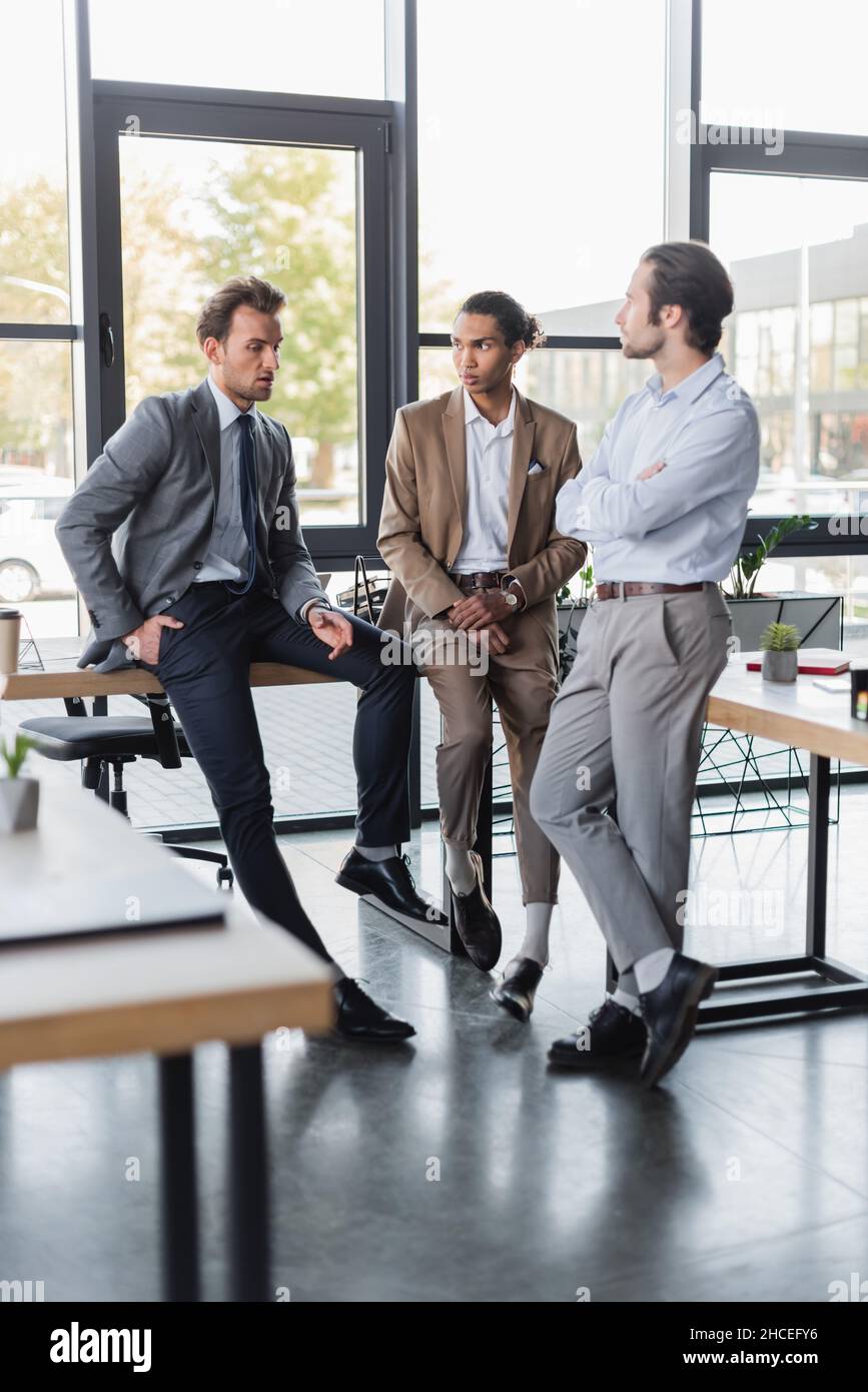 multicultural businessmen in formal wear sitting on desks while talking in office Stock Photo ...