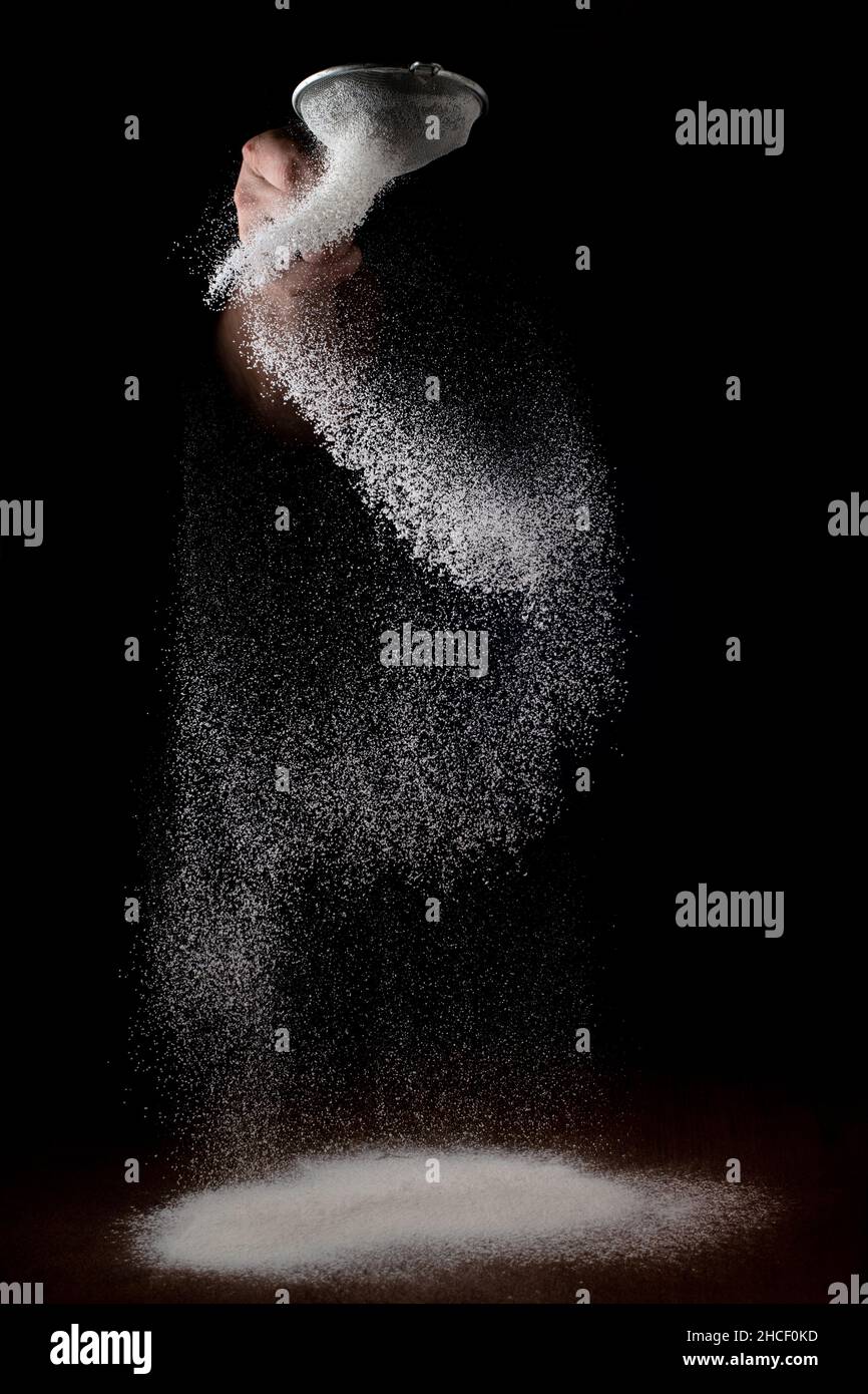 Flour flies through a sieve on a black background. A man sifts flour ...