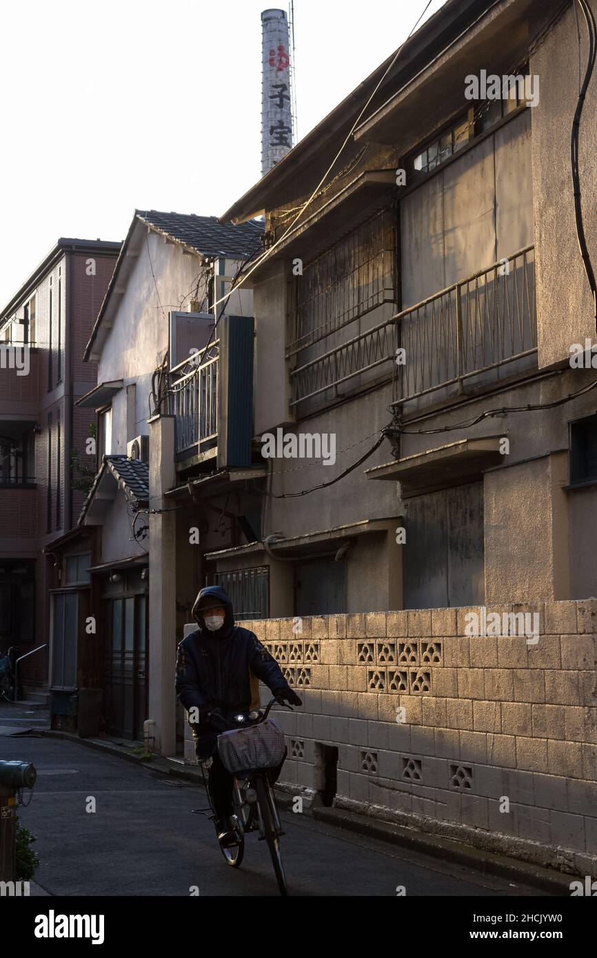 An old man rides a bicycle past the chimney of a Sento or Japanese ...