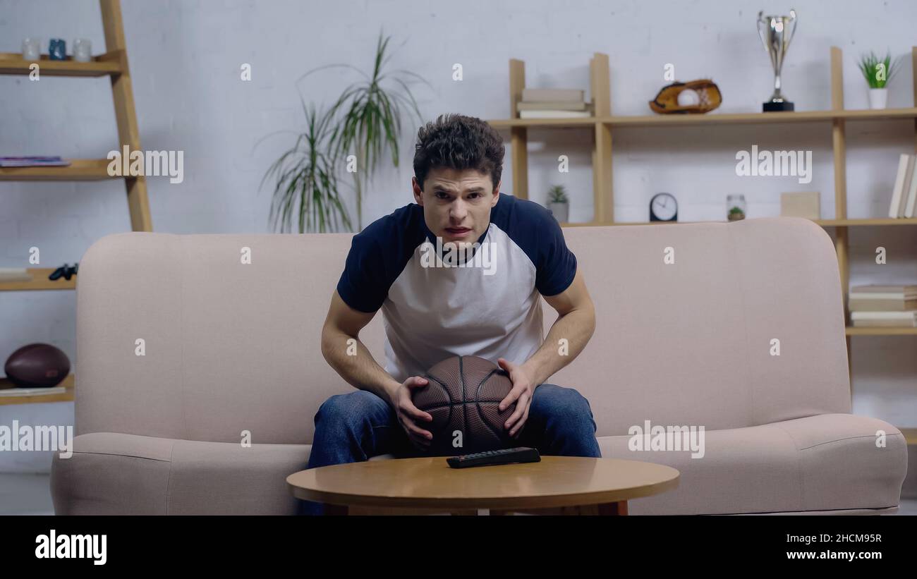worried young man holding ball while watching basketball match on tv at ...