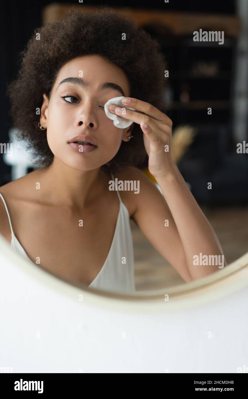 Young african american woman cleaning eye with cotton pad near mirror ...