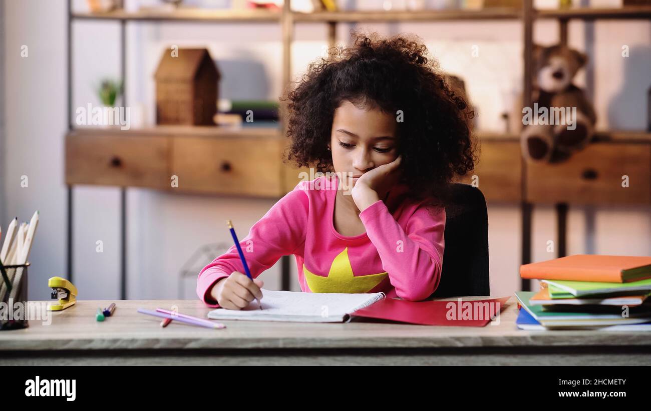 bored african american girl writing in notebook while doing homework ...