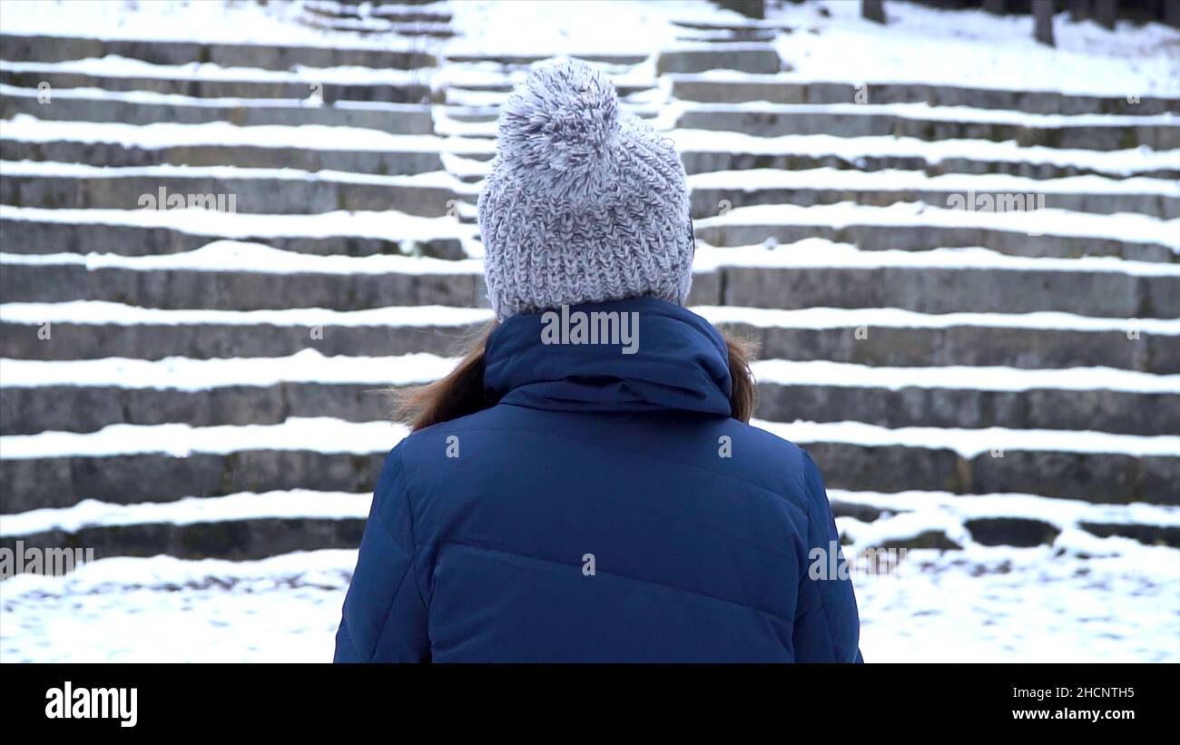 Rear view on woman with hat stays in a park while snowing, snowy stairs ...