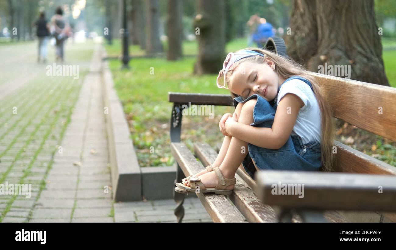 Small tired child girl sitting on a bench with closed eyes resting in ...
