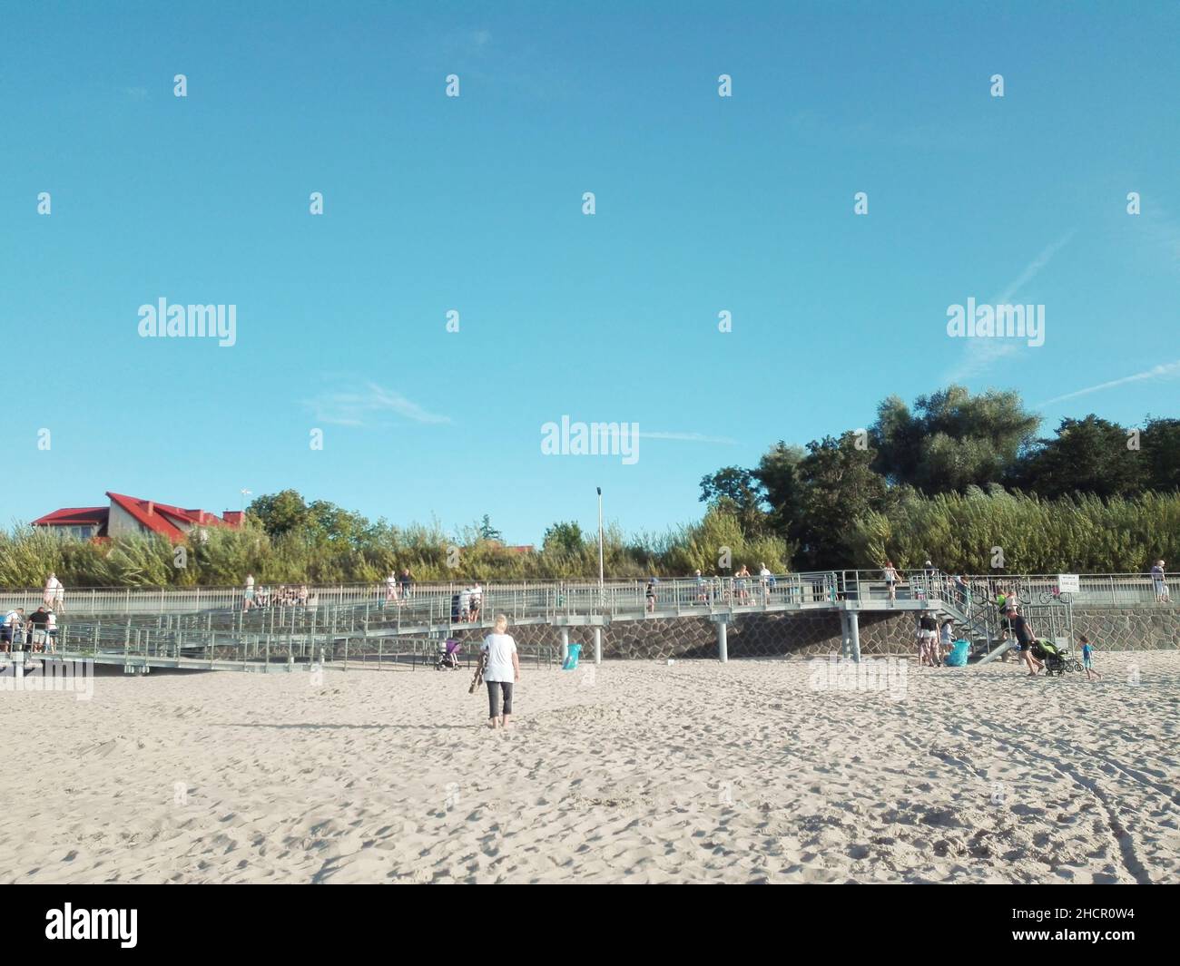 People walking on a metal grid way by a beach against green trees in ...