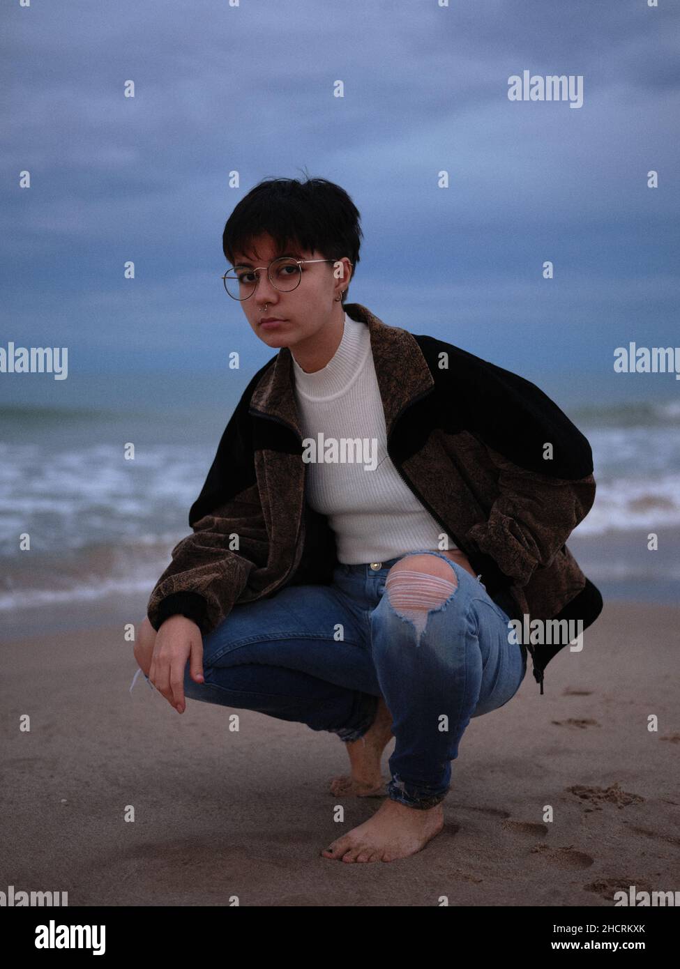 woman squatting barefoot posing on the seashore wearing long trousers ...