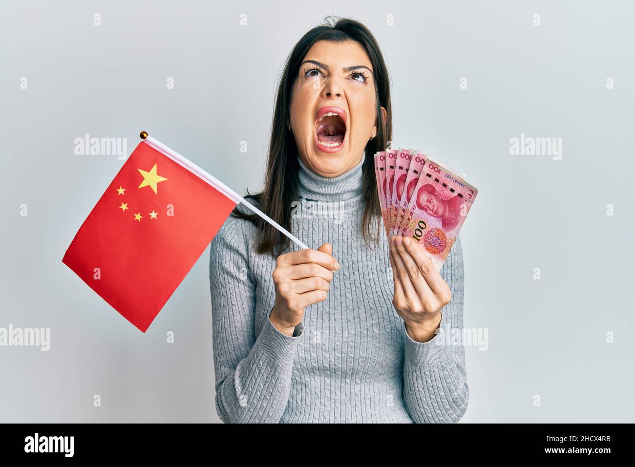 Young hispanic woman holding china flag and yuan banknotes angry and ...