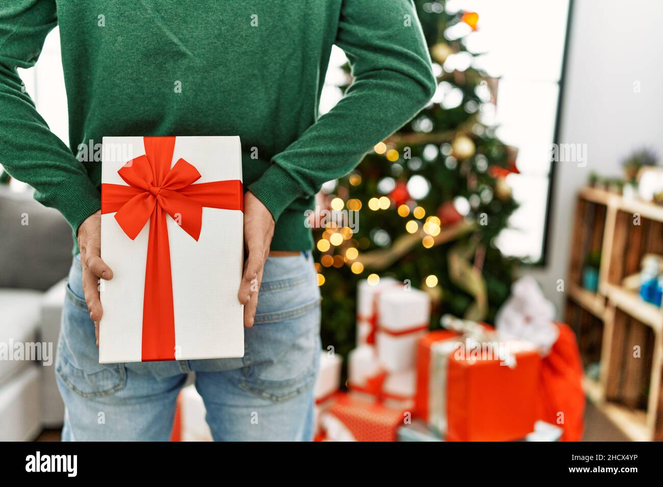 Young redhead man holding gift on back standing by christmas tree at ...