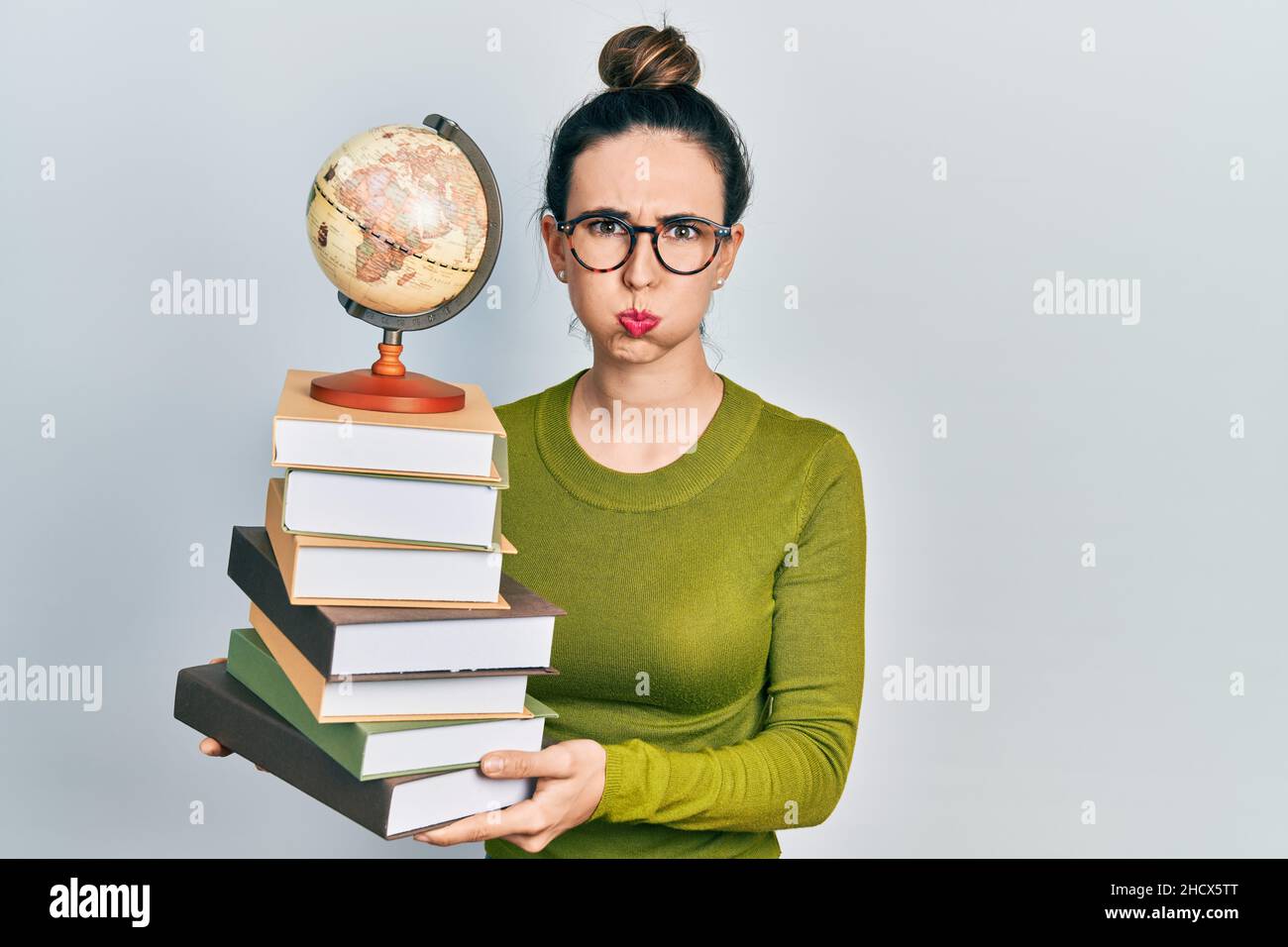Young hispanic girl holding a pile of books and world ball puffing cheeks with funny face. mouth ...