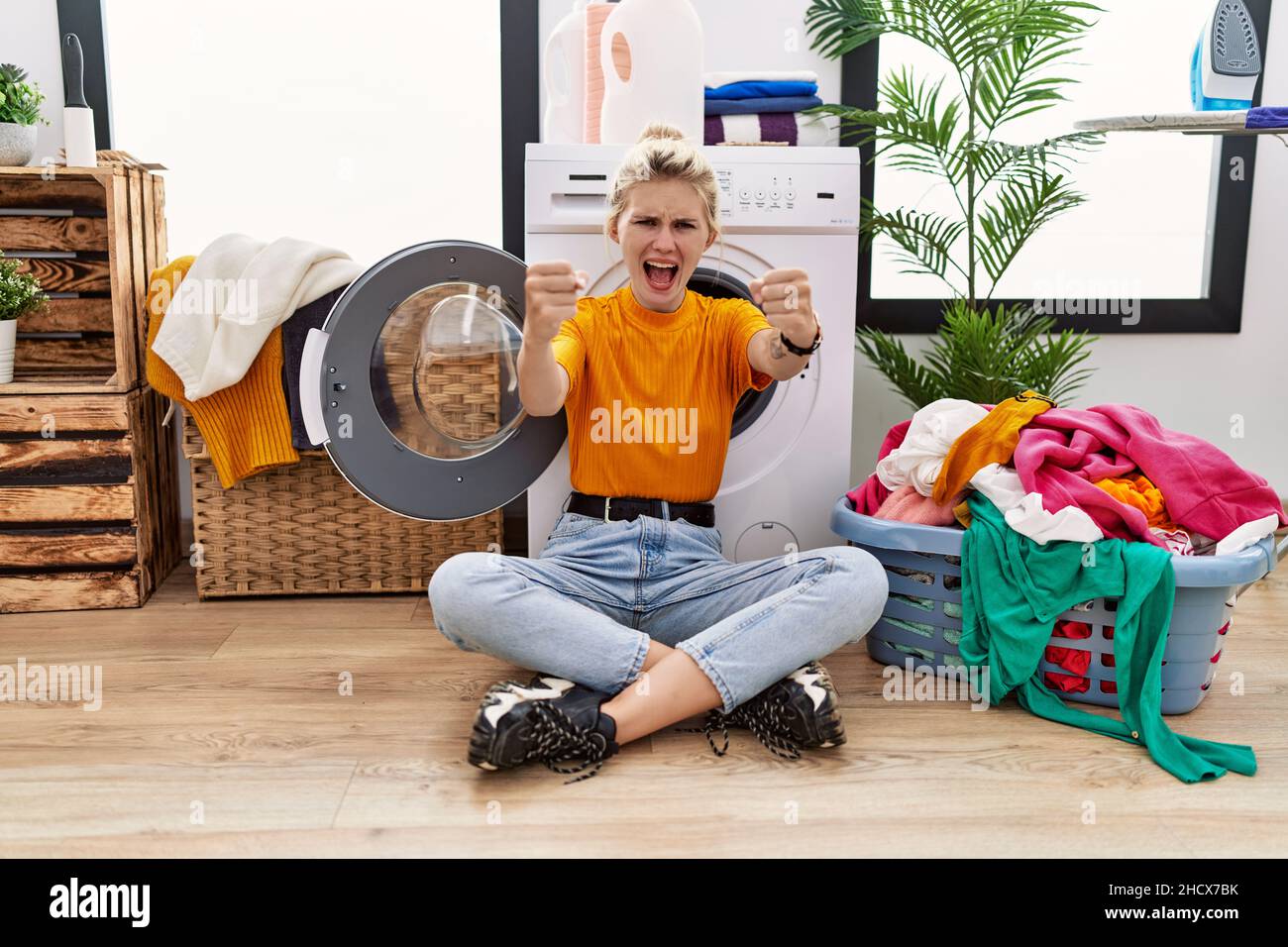 Young blonde woman doing laundry sitting by washing machine angry and ...