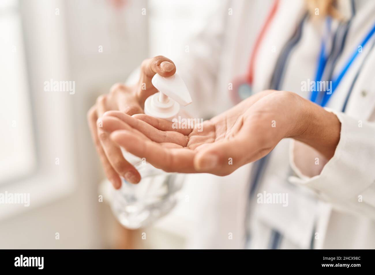 Young blonde woman wearing doctor uniform using sanitizer gel hands at ...