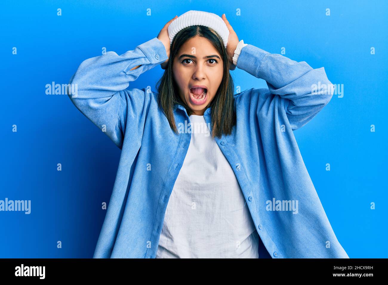 Young latin woman wearing cute wool cap crazy and scared with hands on ...