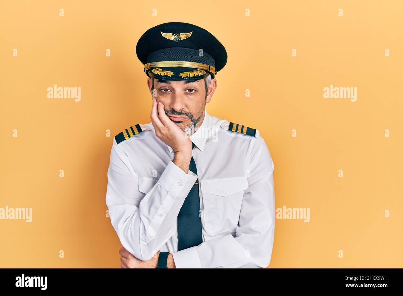 Handsome middle age man with grey hair wearing airplane pilot uniform thinking looking tired and ...