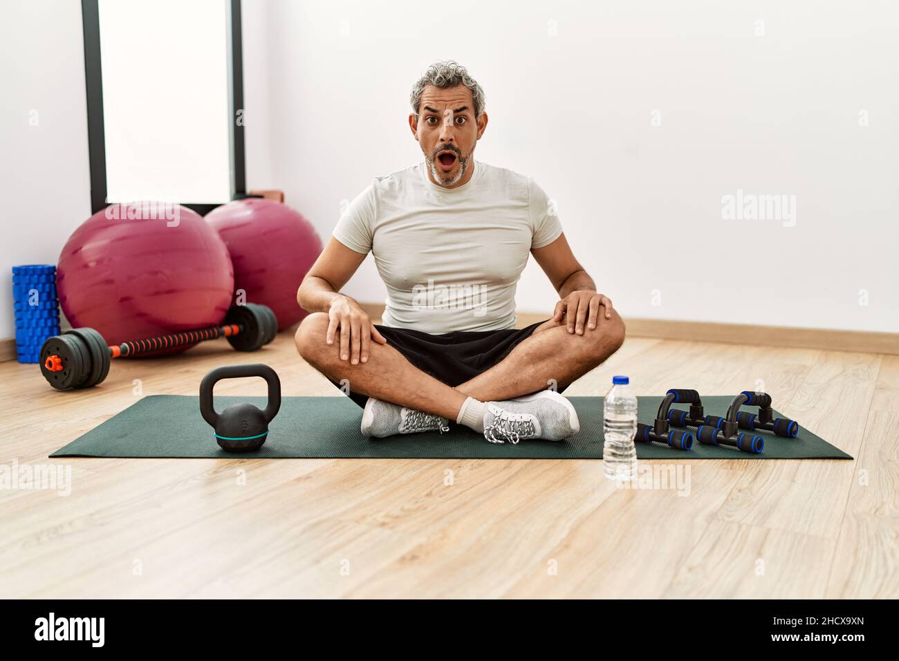 Middle age hispanic man sitting on training mat at the gym scared and ...