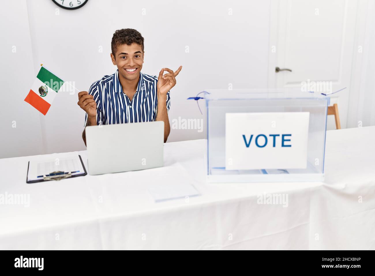 Young handsome hispanic man at political campaign election holding mexico flag smiling happy ...