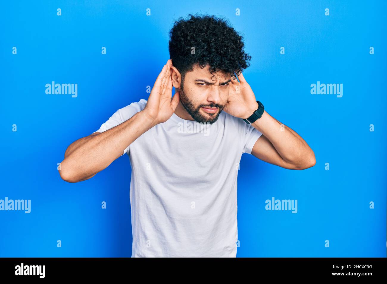 Young arab man with beard wearing casual white t shirt trying to hear ...