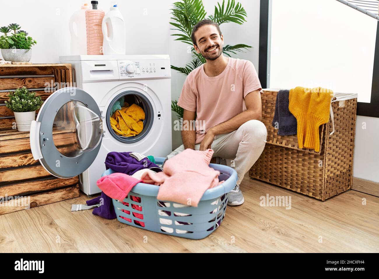 Young hispanic man putting dirty laundry into washing machine looking ...