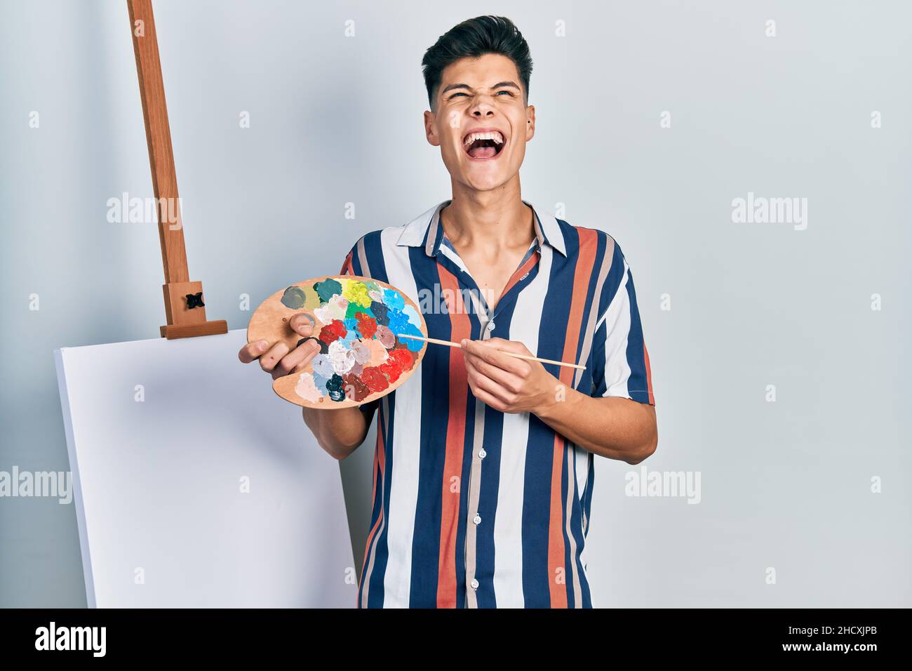 Young hispanic man holding painter palette and paintbrush standing ...