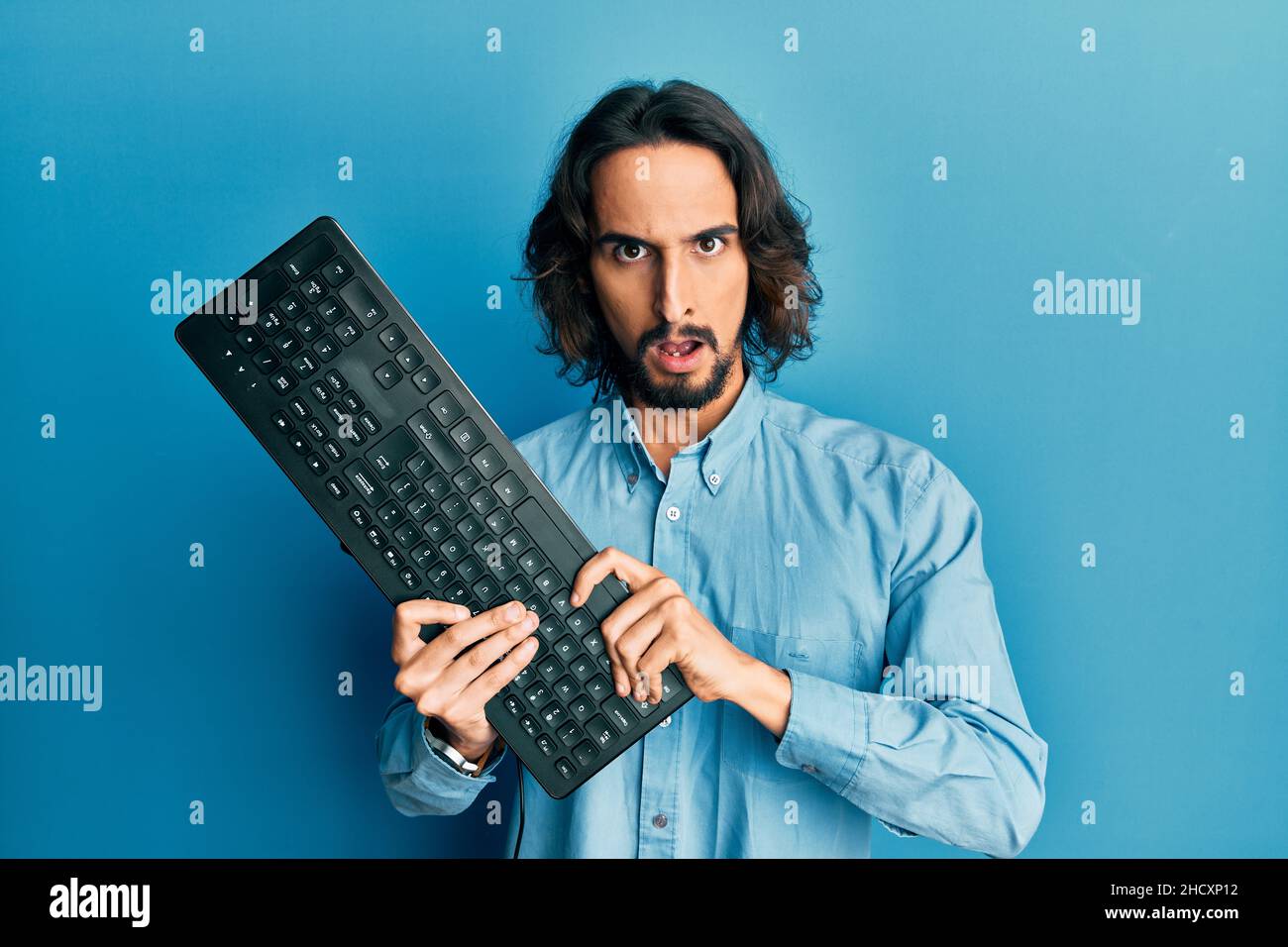 Young hispanic man holding keyboard in shock face, looking skeptical ...