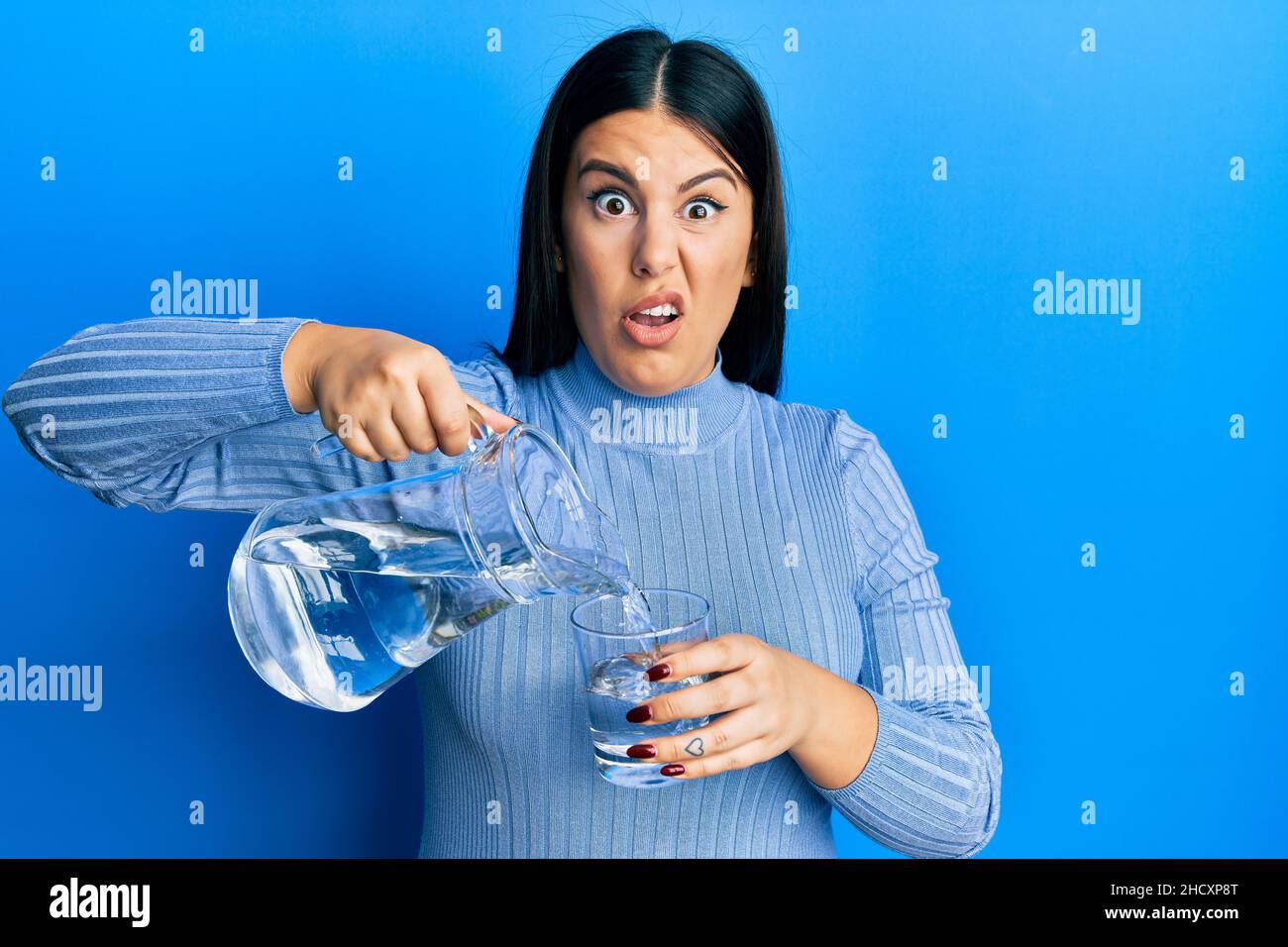 Beautiful brunette woman pouring water in glass clueless and confused ...