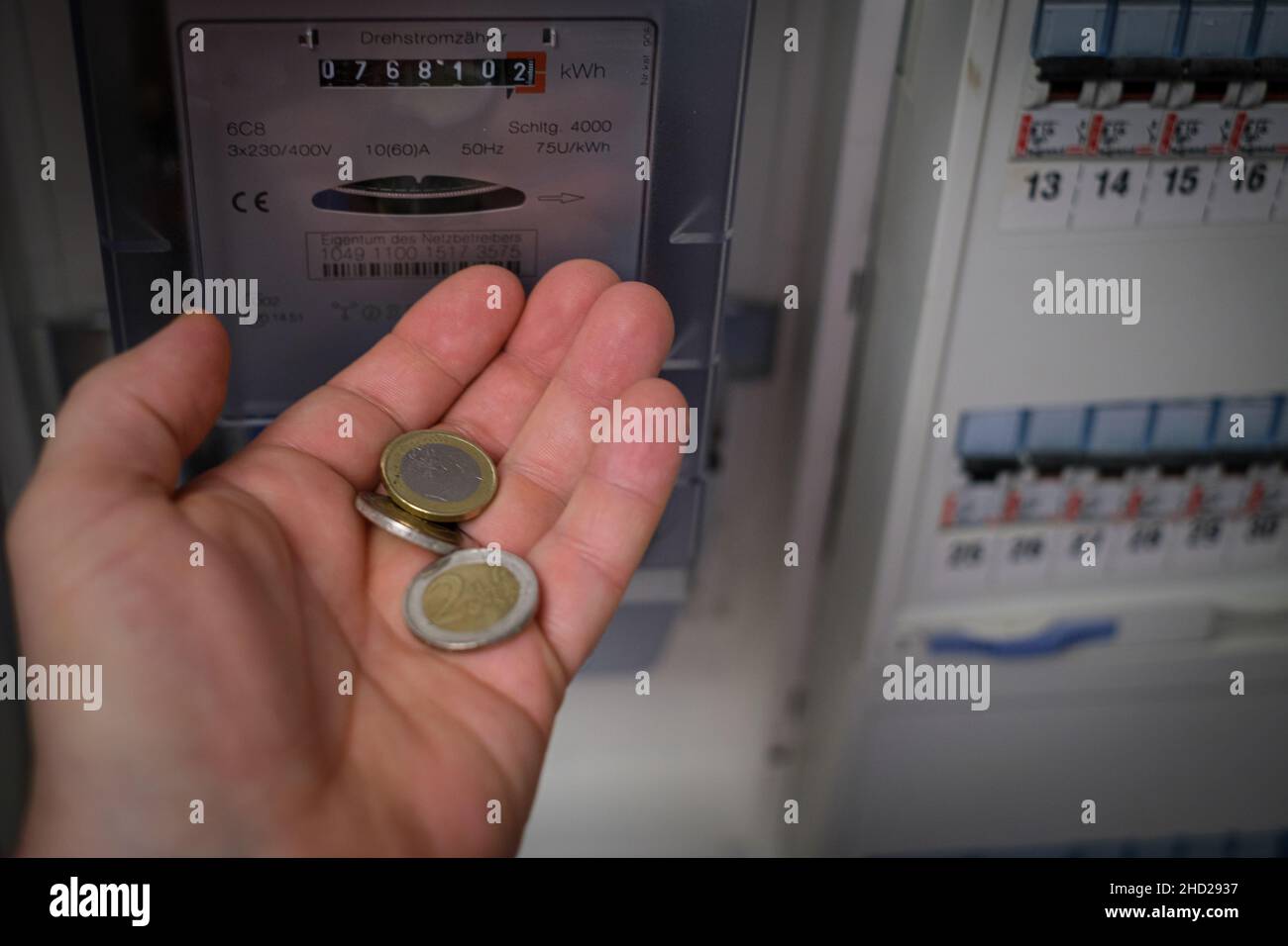 in one hand are several euro coins in front of a three-phase meter ...