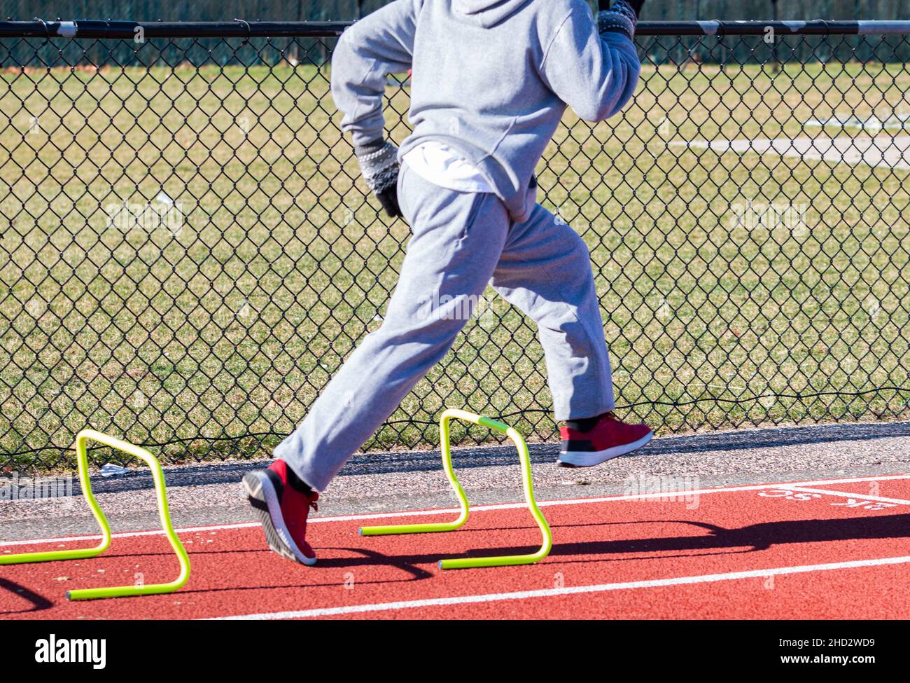 A high school runner wearing a gray sweatsuit is running over yellow mini hurdles on a red track ...
